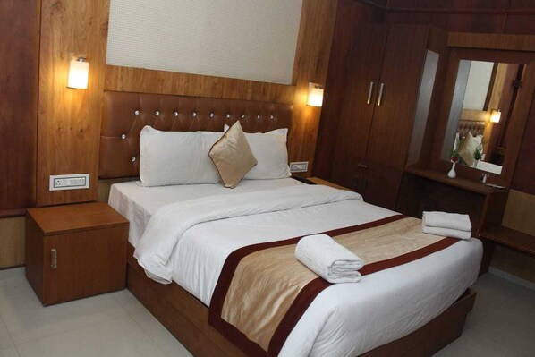 Cama bem arrumada com roupa de cama branca e detalhes em castanho, mesa de cabeceira de madeira, guarda-roupa de madeira e um espelho sobre a cama.
