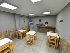 Quarto com seis mesas e cadeiras de madeira, cozinha compacta equipada e uma janela ampla que proporciona iluminação natural.