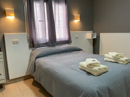 Quarto com cama, duas luminárias fixadas na parede, janela com cortinas e duas toalhas dobradas sobre a cama.
