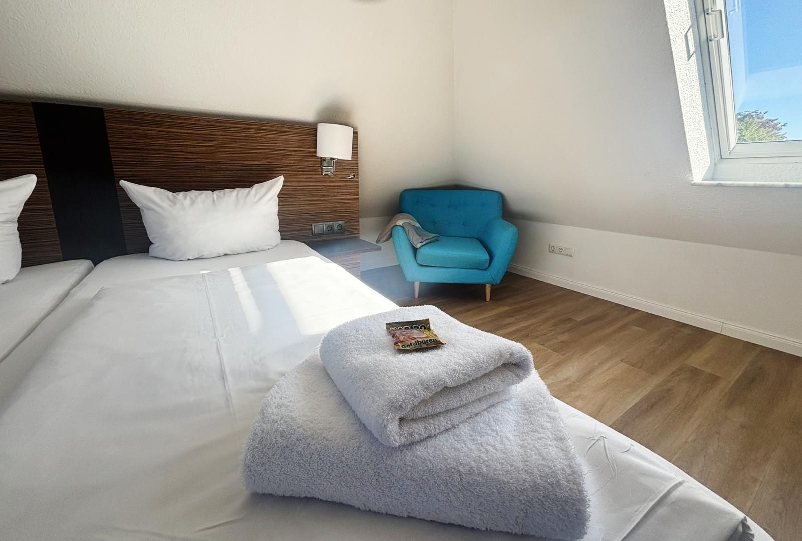 Quarto de hotel com uma cama grande, uma poltrona azul e uma janela.