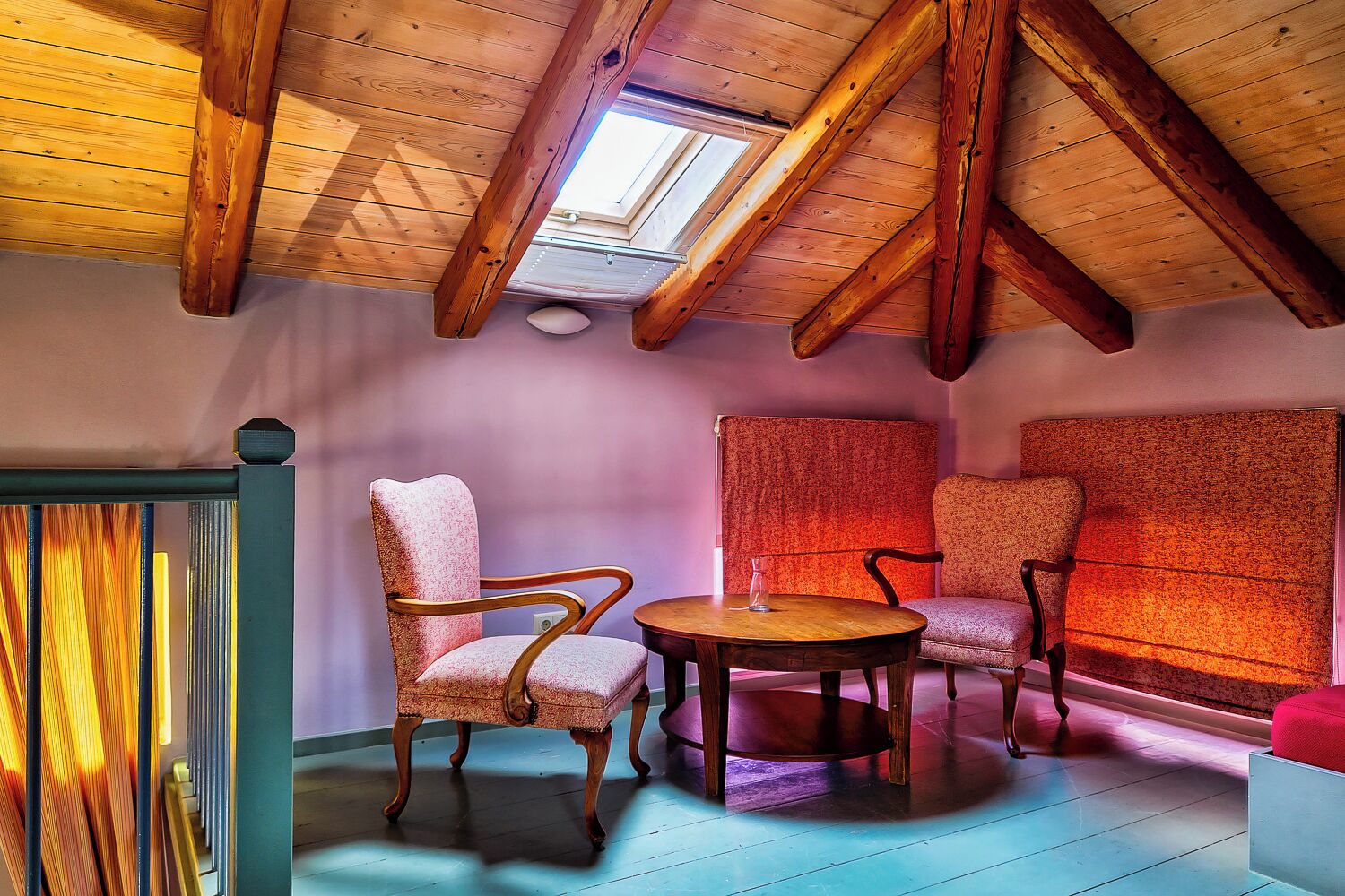 Um quarto de sótão acolhedor, com vigas de madeira, um claraboia e duas poltronas estofadas ao redor de uma mesa pequena.