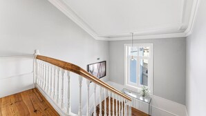 Uma escadaria com grades brancas e degraus de madeira, que leva a um quarto com janela e um quadro na parede.