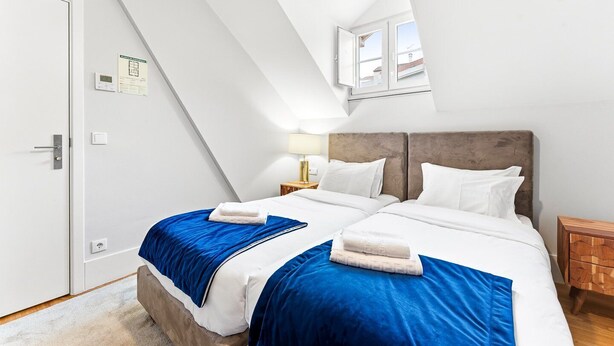 Quarto com teto inclinado, cama com roupa de cama azul e branca, mesa de cabeceira e janela com vista para edifícios.