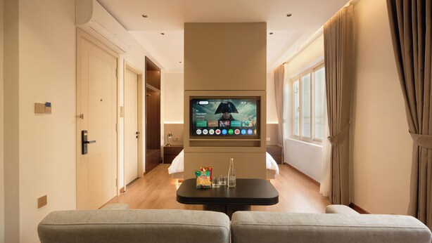 Quarto de hotel moderno com televisão de tela plana em suporte, sofá, mesinha com petiscos e janela com cortinas.