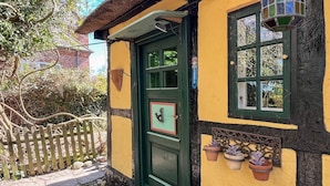 Uma casa tradicional de madeira e tijolo com uma porta verde, uma pequena janela com vitrais e plantas em vasos na parede.