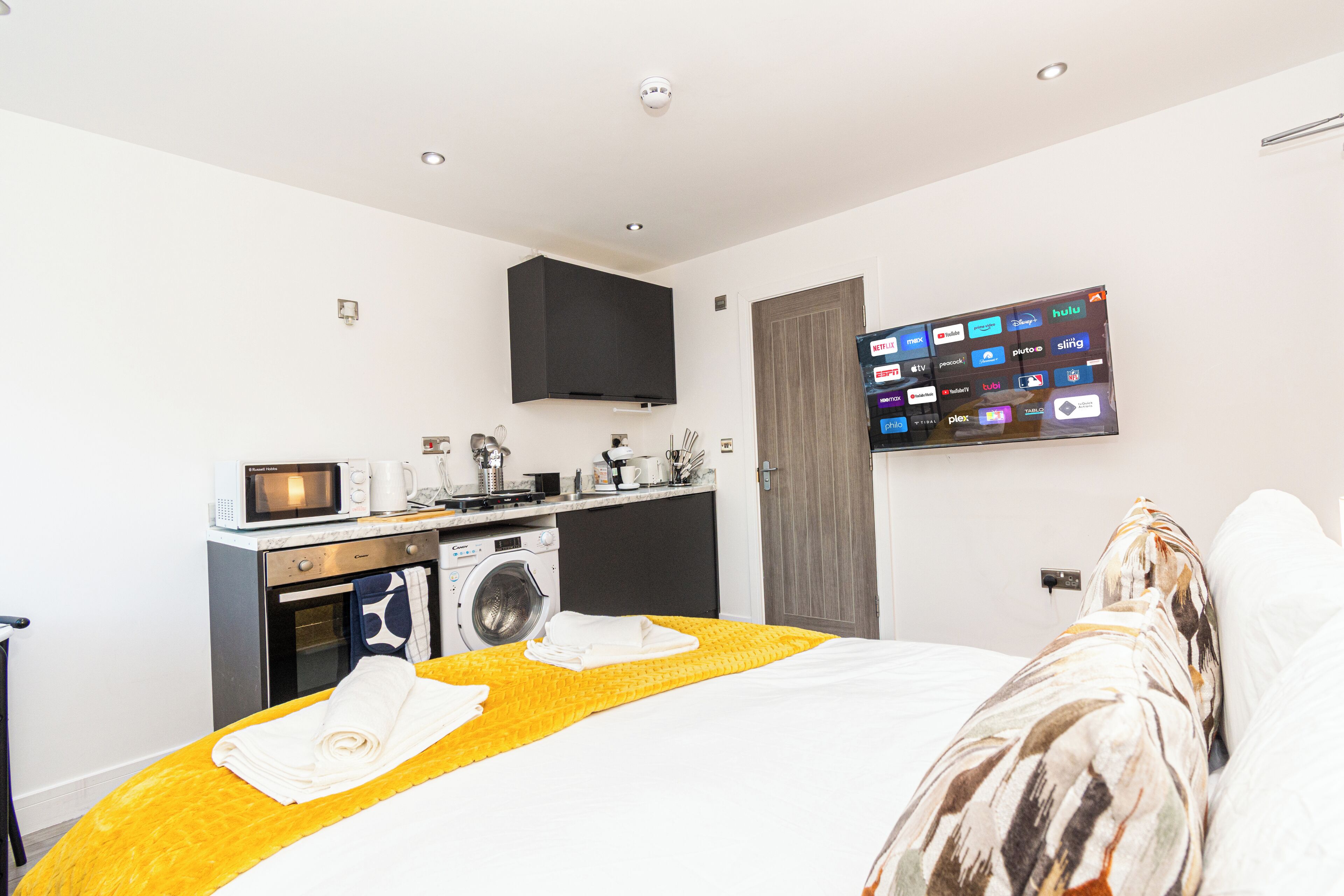 Quarto de hotel com cama, televisão de tela plana exibindo serviços de streaming, micro-ondas, cafeteira e máquina de lavar.