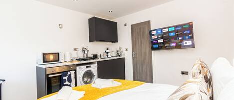 Quarto de hotel com cama, televisão de tela plana exibindo serviços de streaming, micro-ondas, cafeteira e máquina de lavar.