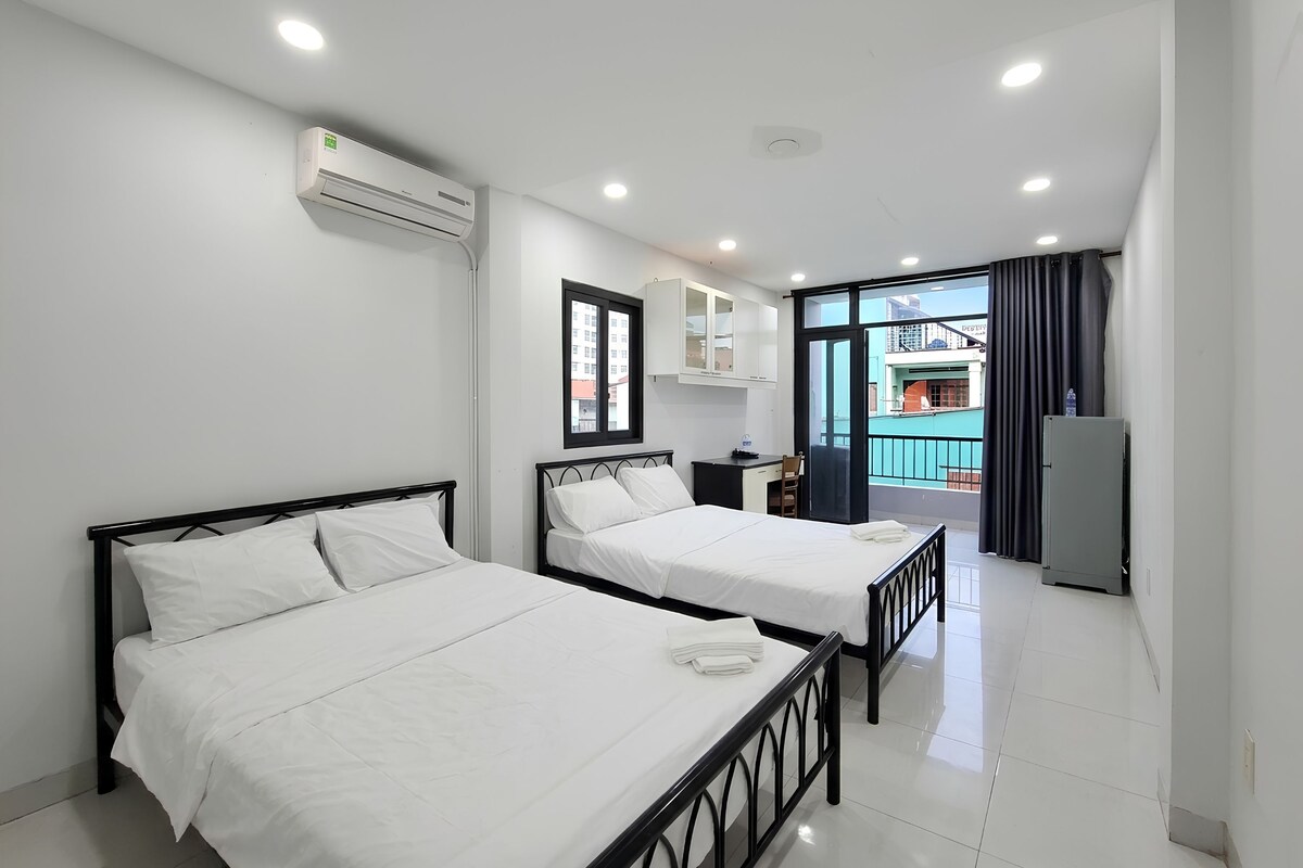 Quarto de hotel com duas camas, uma cozinha compacta e varanda com vista para edifícios.