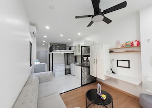 Quarto moderno e bem iluminado, com um sofá branco, um ventilador de teto preto e uma pequena mesa redonda com um brinquedo.