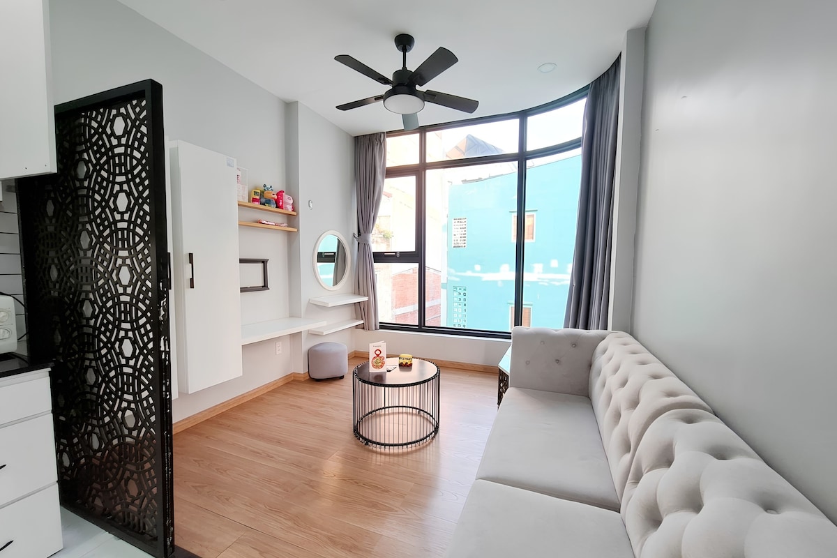 Uma sala de estar moderna com sofá, mesa de centro redonda, ventilador de teto e uma grande janela com cortinas.