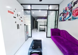Quarto de hotel moderno com sofá roxo, mesa de centro de vidro, cama com cabeceira branca e uma janela grande.