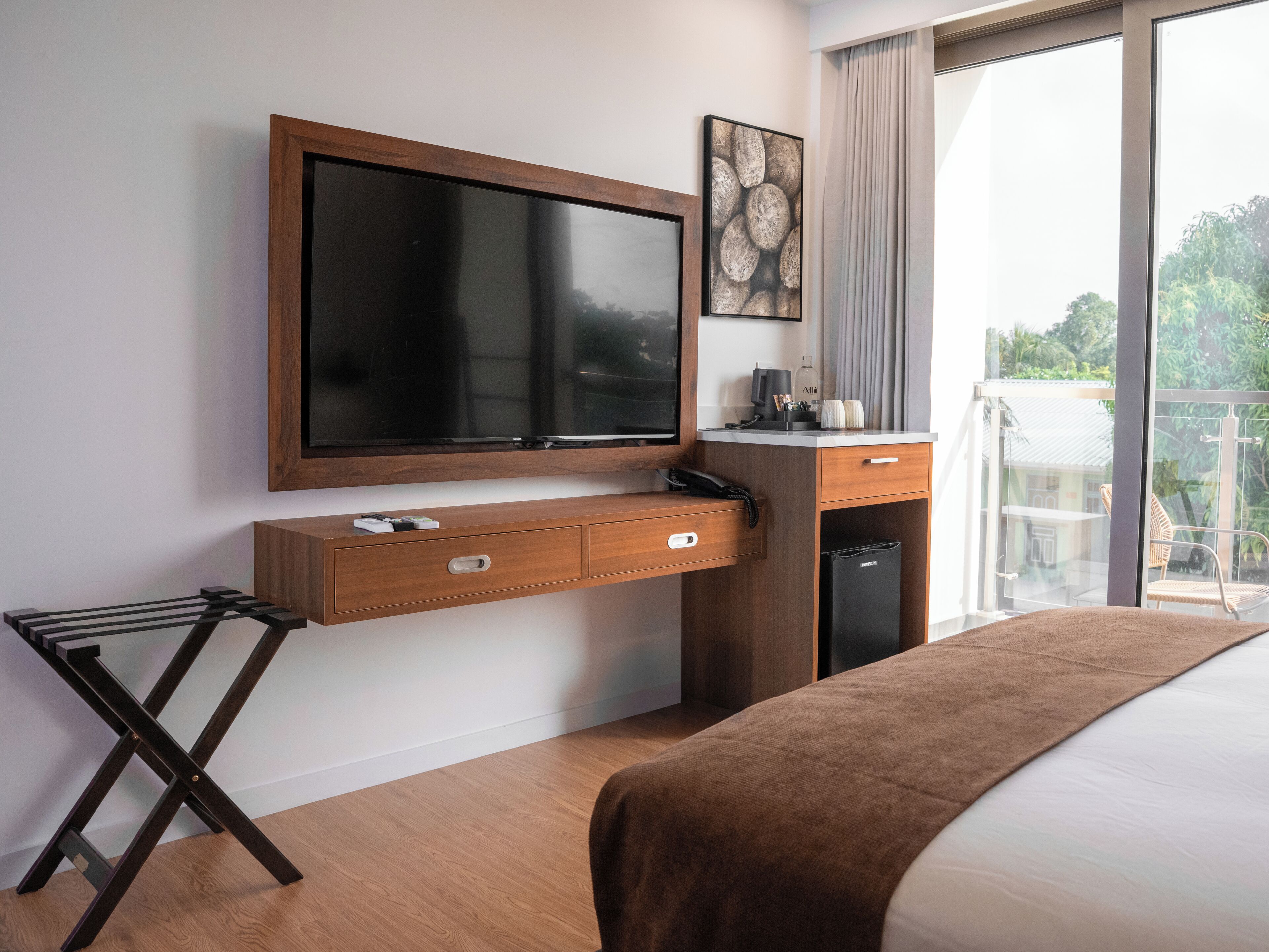 Quarto de hotel com uma televisão de tela plana grande na parede, uma escrivaninha de madeira com telefone e uma máquina de café pequena, uma geladeira preta compacta e uma cama com um cobertor marrom.