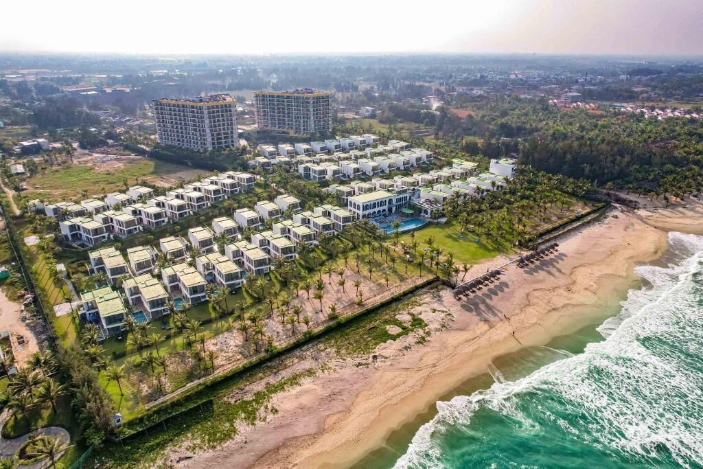 Vista aérea de um resort costeiro com vários edifícios tipo bangalô, uma praia e ondas do oceano.