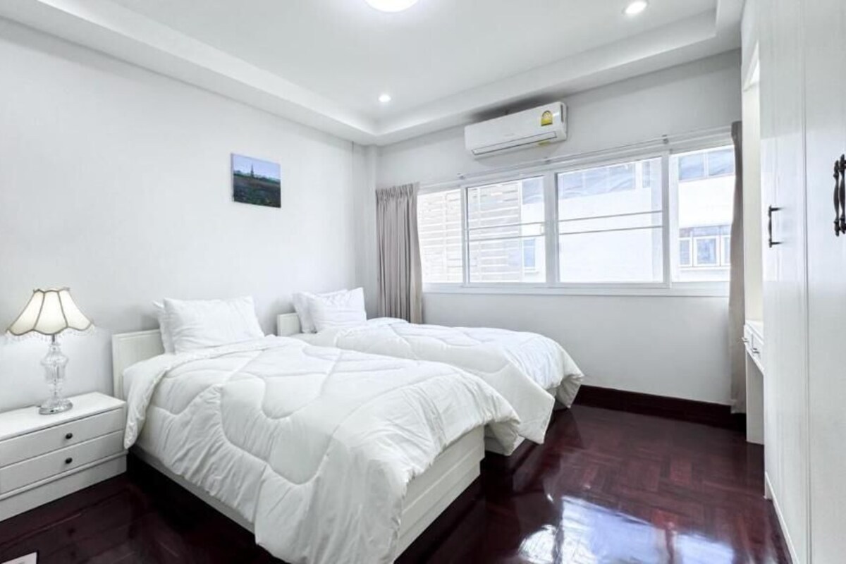 Quarto com cama, mesa de cabeceira, luminária e janela com cortinas.