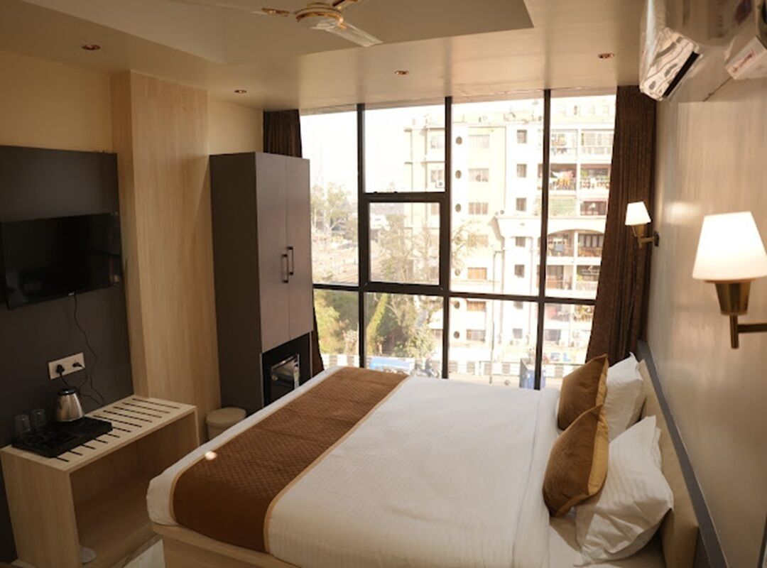 Quarto de hotel com uma cama grande, uma mesa com chaleira, televisão e janela com vista para edifícios.