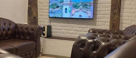 Sala de estar com parede de tijolos brancos, TV montada e sofás de couro marrom.