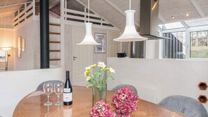 Uma sala de jantar com mesa de madeira, luminárias pendentes brancas, uma garrafa de vinho e um vaso de flores.