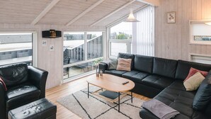 Uma sala de estar moderna com um sofá de couro preto, uma mesa de centro de madeira e uma poltrona preta. Há uma janela grande que oferece vista para o exterior.