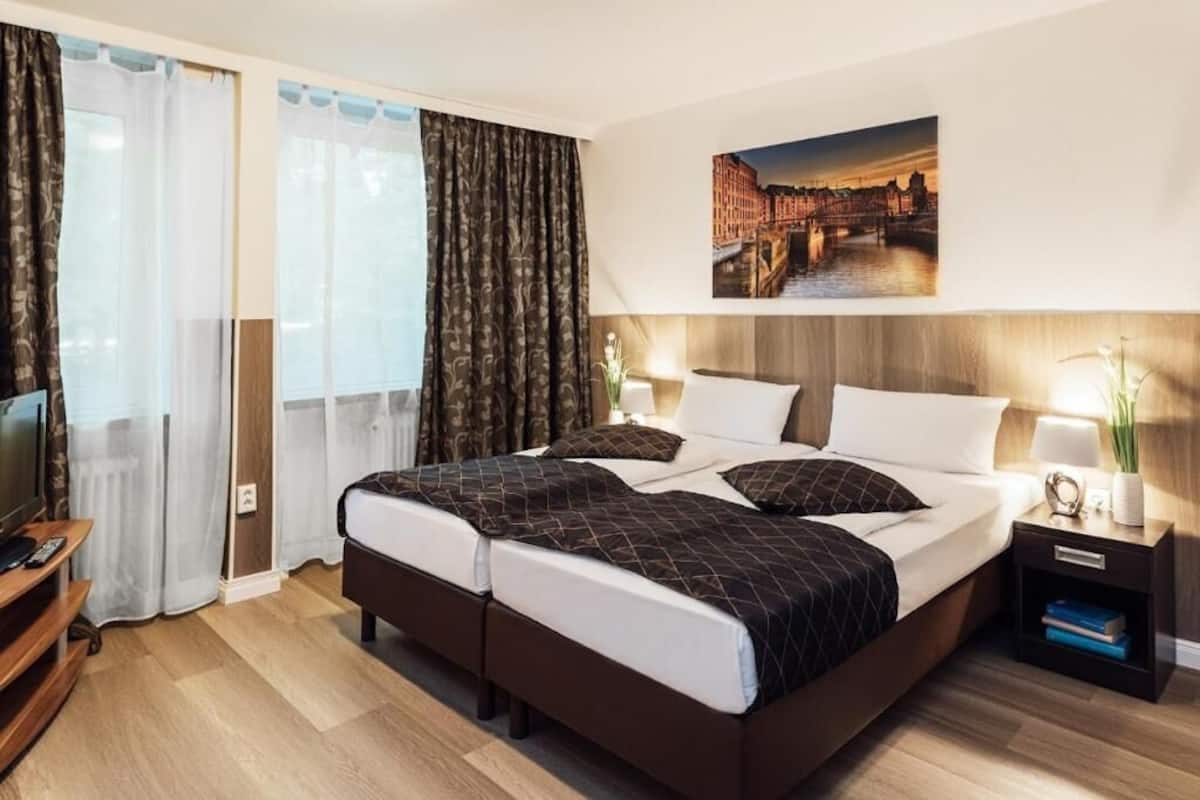Uma cama de casal com cabeceira escura, uma mesa de cabeceira com abajur, e uma pintura com vista para a cidade na parede.