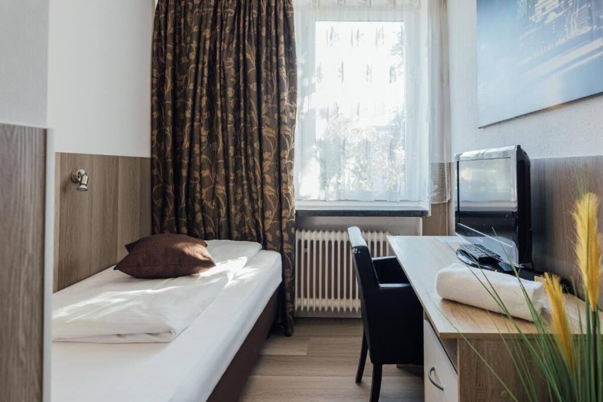 Quarto de hotel com cama, escrivaninha, cadeira, televisão e janela com cortinas.