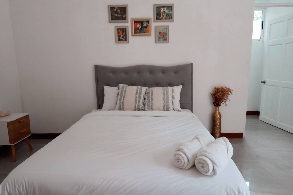 Uma cama bem arrumada com cabeceira cinzenta, uma mesinha de cabeceira com gaveta e um vaso com flores secas no criado-mudo.