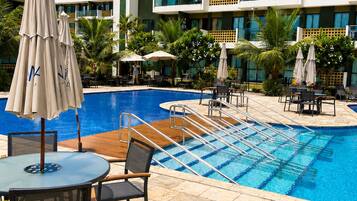 Uma área de piscina de hotel com várias espreguiçadeiras e guarda-sóis, cercada por um edifício com vários andares e varandas.