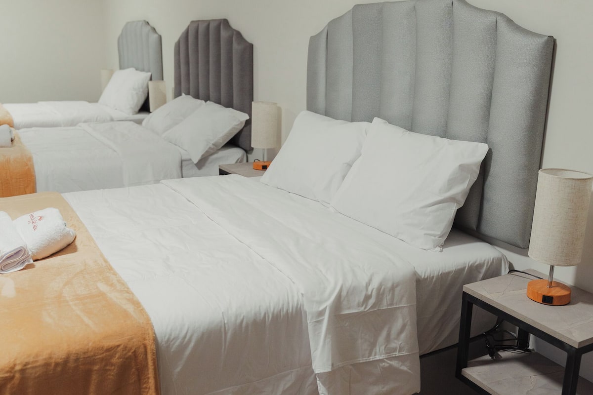 Quarto de hotel com duas camas, cada uma com cabeceira cinza, roupa de cama branca e uma pequena mesa de cabeceira com abajur.