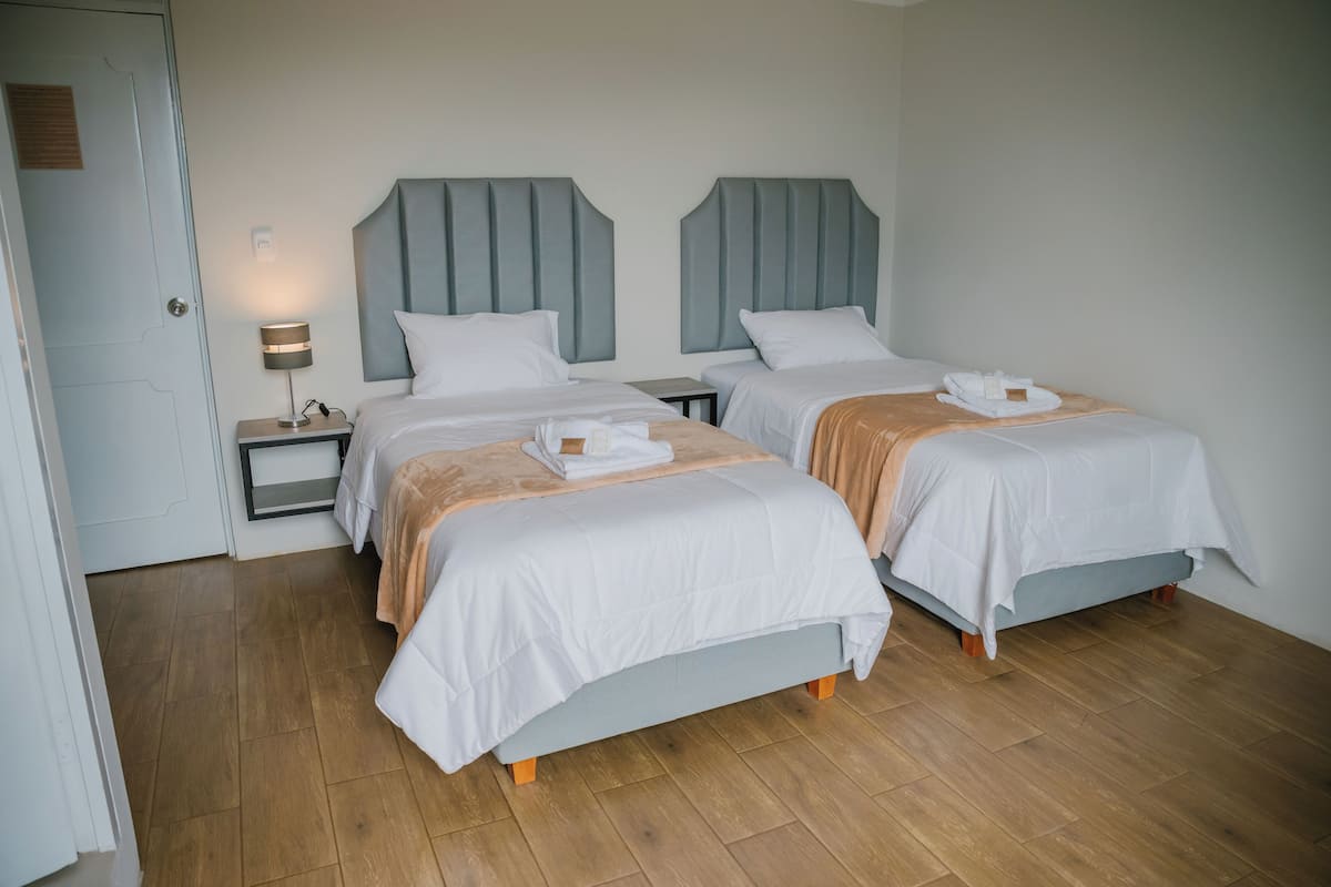 Quarto de hotel com duas camas, uma mesa de cabeceira com abajur e uma porta.