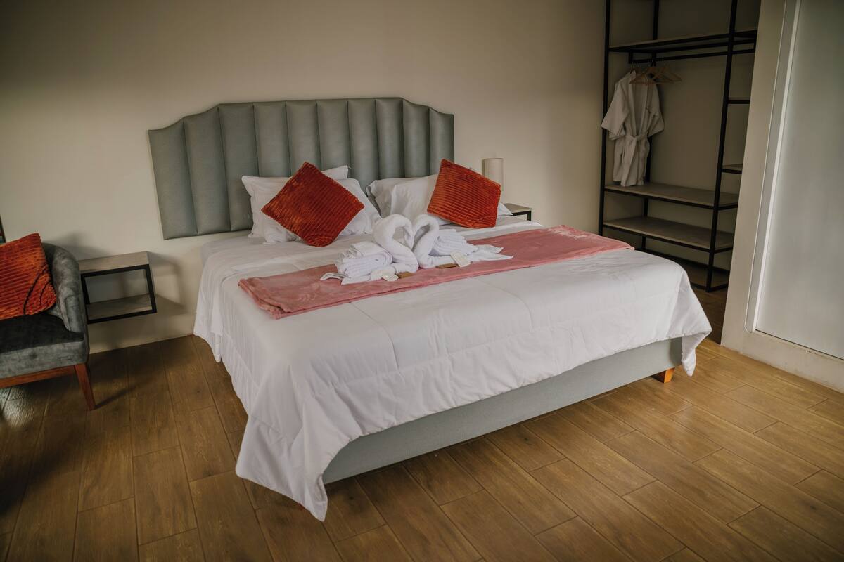 Uma cama bem arrumada com lençóis brancos e almofadas vermelhas, uma cabeceira estofada cinza e piso de madeira.