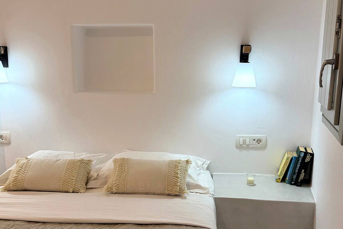 Quarto de hotel com uma cama, duas luminárias de cabeceira, mesa de cabeceira com livros e uma pequena mesa com um copo de leite.