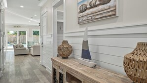 Um corredor com piso de madeira, um sofá branco, uma mesa de jantar com um vaso e uma escultura, e um quadro na parede.