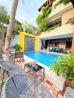 Área de piscina com espreguiçadeiras e uma mesa, rodeada por plantas e uma parede amarela.
