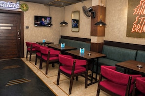 Interior de restaurante com mesas de madeira, cadeiras vermelhas, um letreiro de neon e uma televisão na parede.