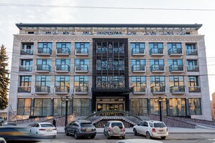 Um hotel com vários andares, varandas, entrada central e estacionamento com diversos carros.