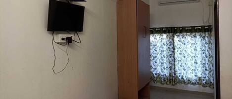 Quarto com cama, televisão fixada na parede, guarda-roupa e janela com cortinas florais.