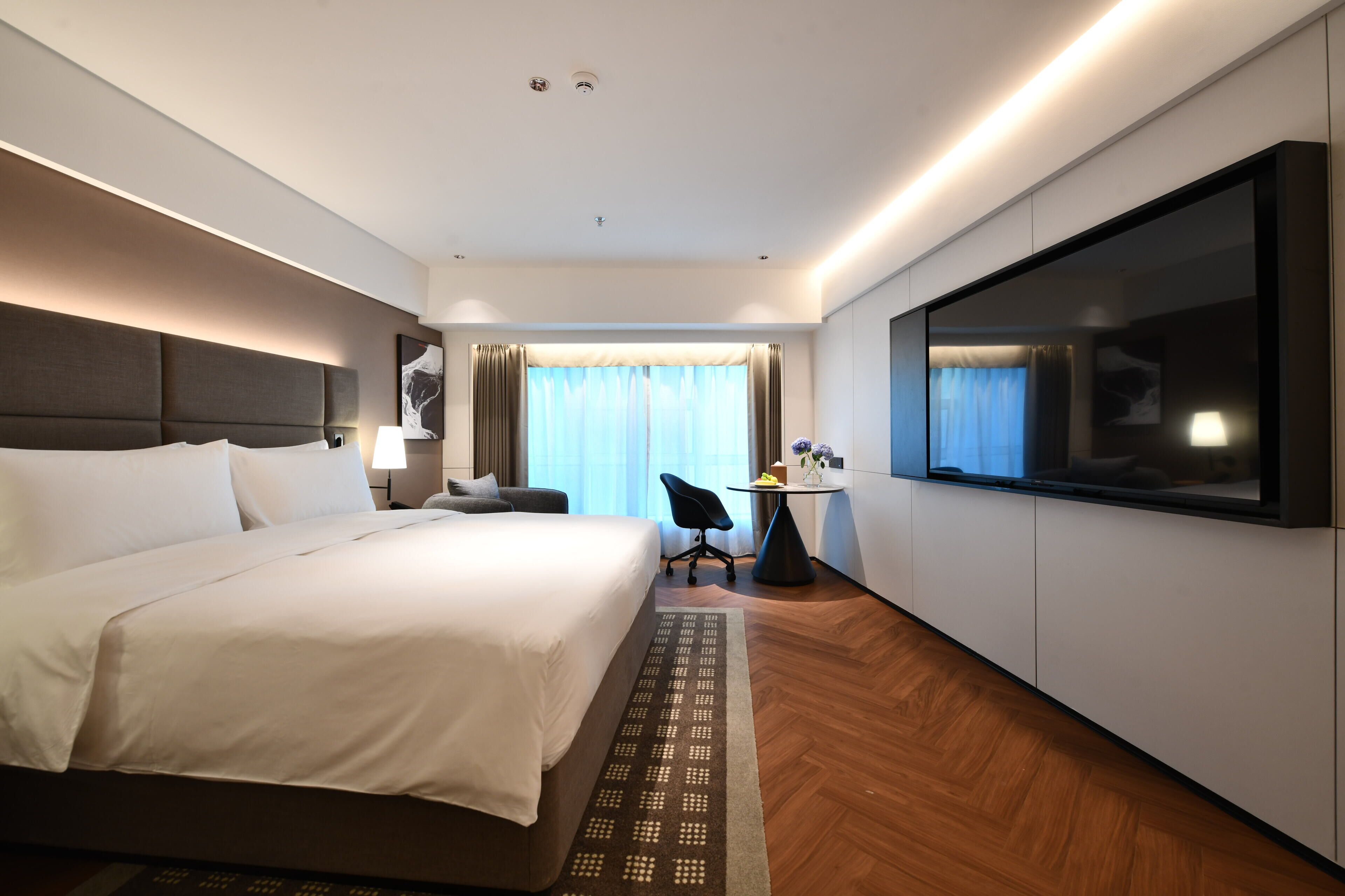 Quarto de hotel moderno com uma cama grande, uma escrivaninha, uma cadeira, uma televisão e uma janela com cortinas.