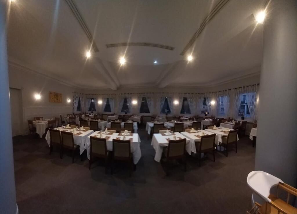 Uma grande sala de banquetes com várias mesas redondas postas para refeições, tetos altos e janelas amplas com cortinas.