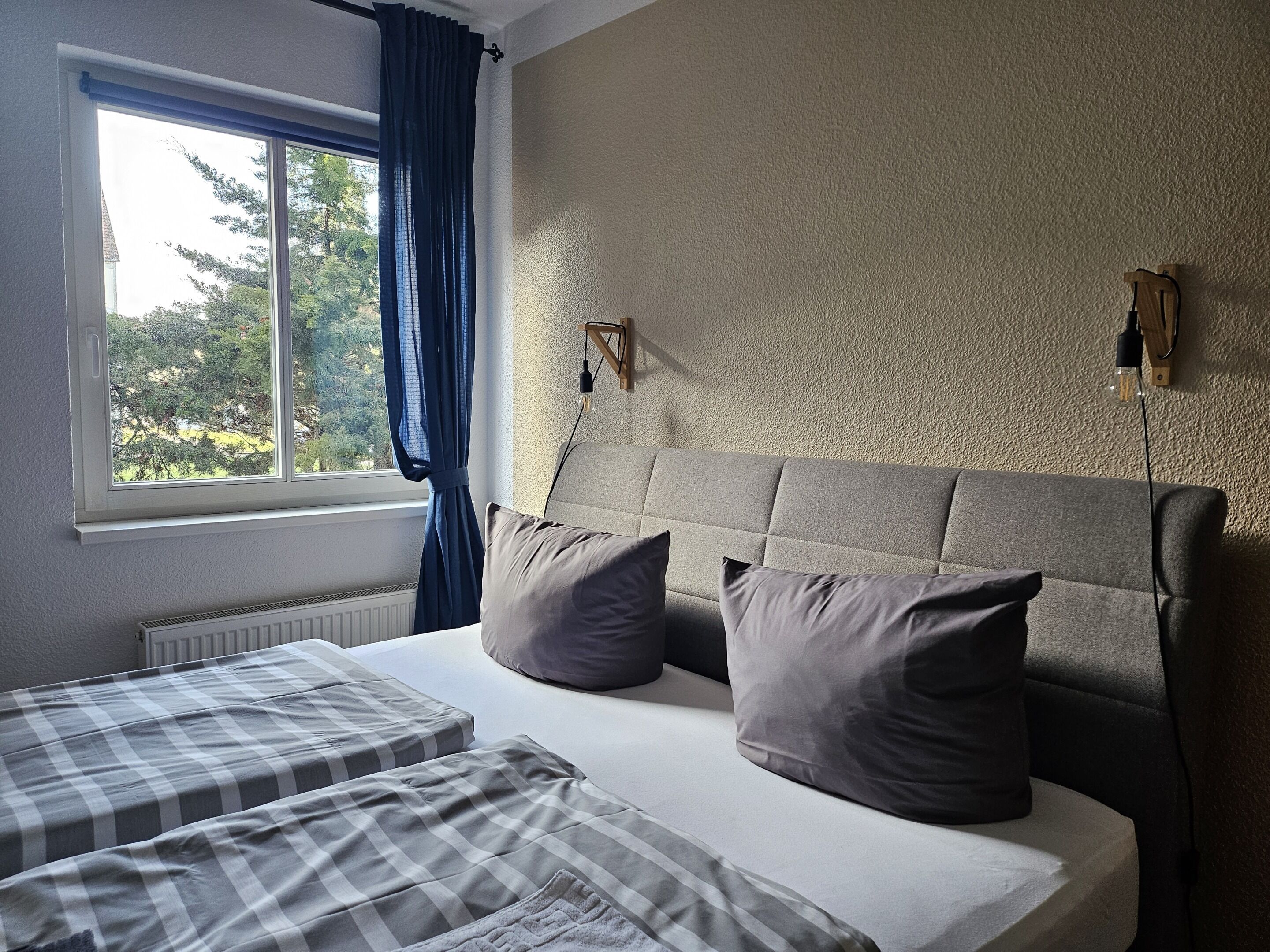 Quarto com cama, duas almofadas, janela com cortinas e luminária fixada na parede.