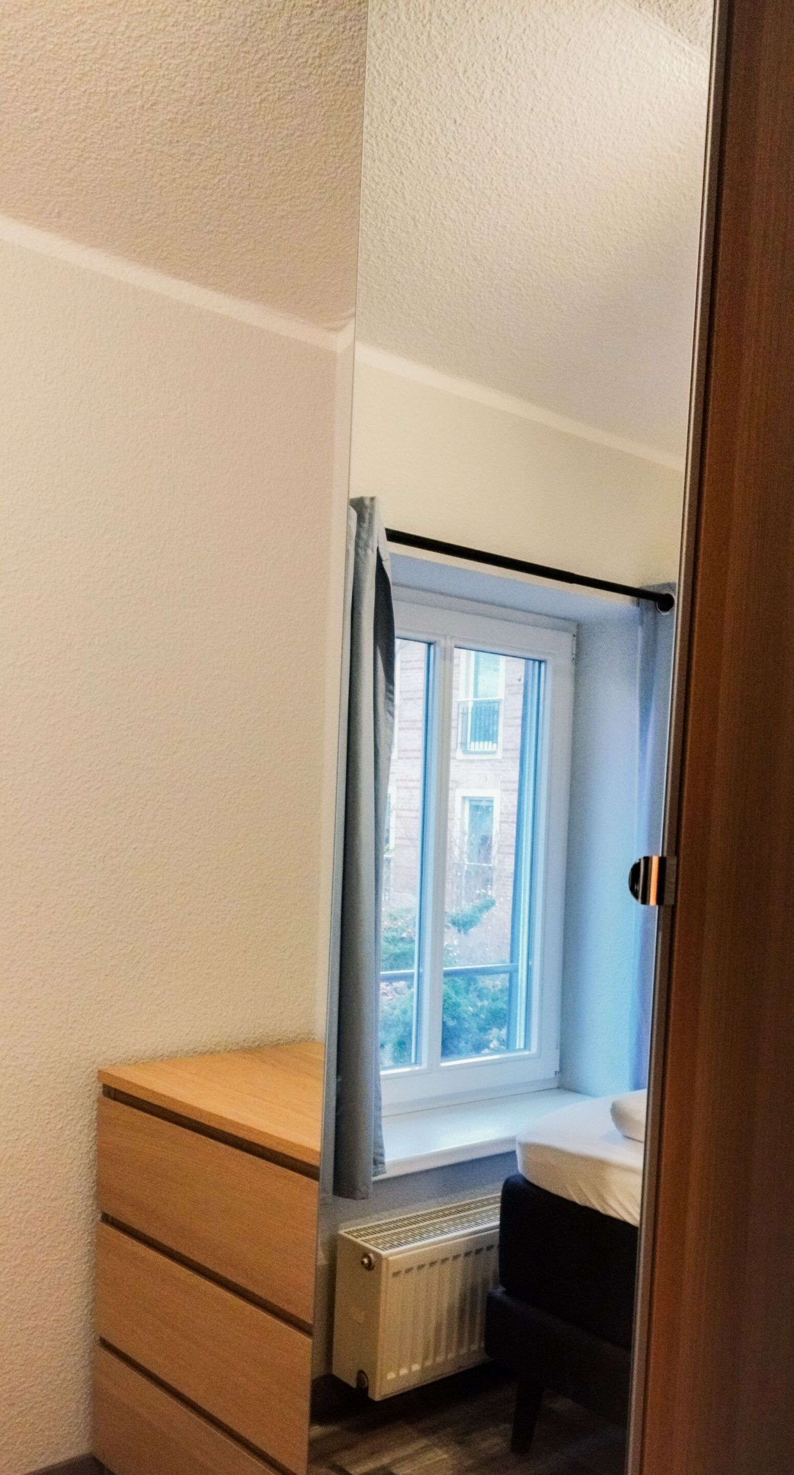 Um quarto pequeno com janela, radiador e um armário de madeira.