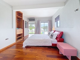 Quarto com uma cama grande, um guarda-roupa de madeira, uma mesa de cabeceira rosa e vista para o exterior através de uma porta de correr de vidro.