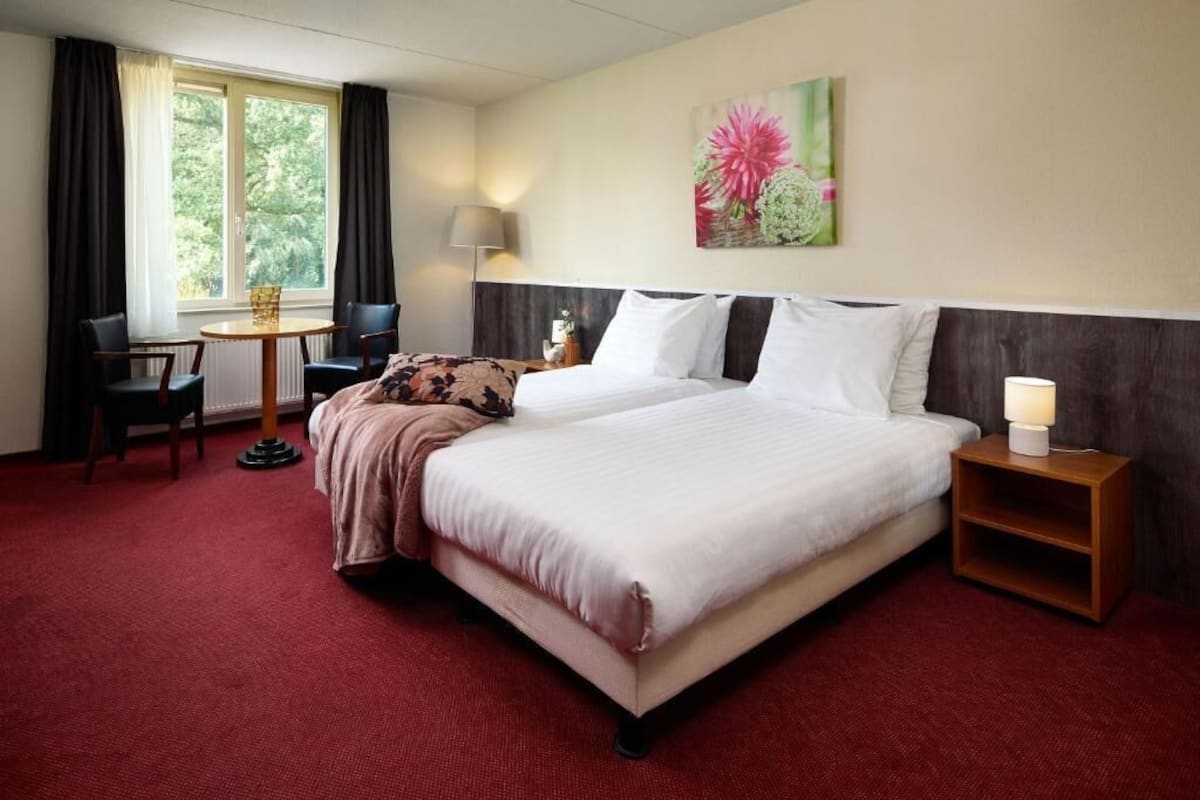 Quarto de hotel com uma cama grande, uma mesa com duas cadeiras, uma luminária e um quadro na parede.