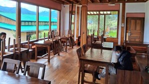 Interior em madeira com grandes janelas que oferecem vista para uma montanha e um telhado turquesa. Mesas e cadeiras dispostas para refeições.
