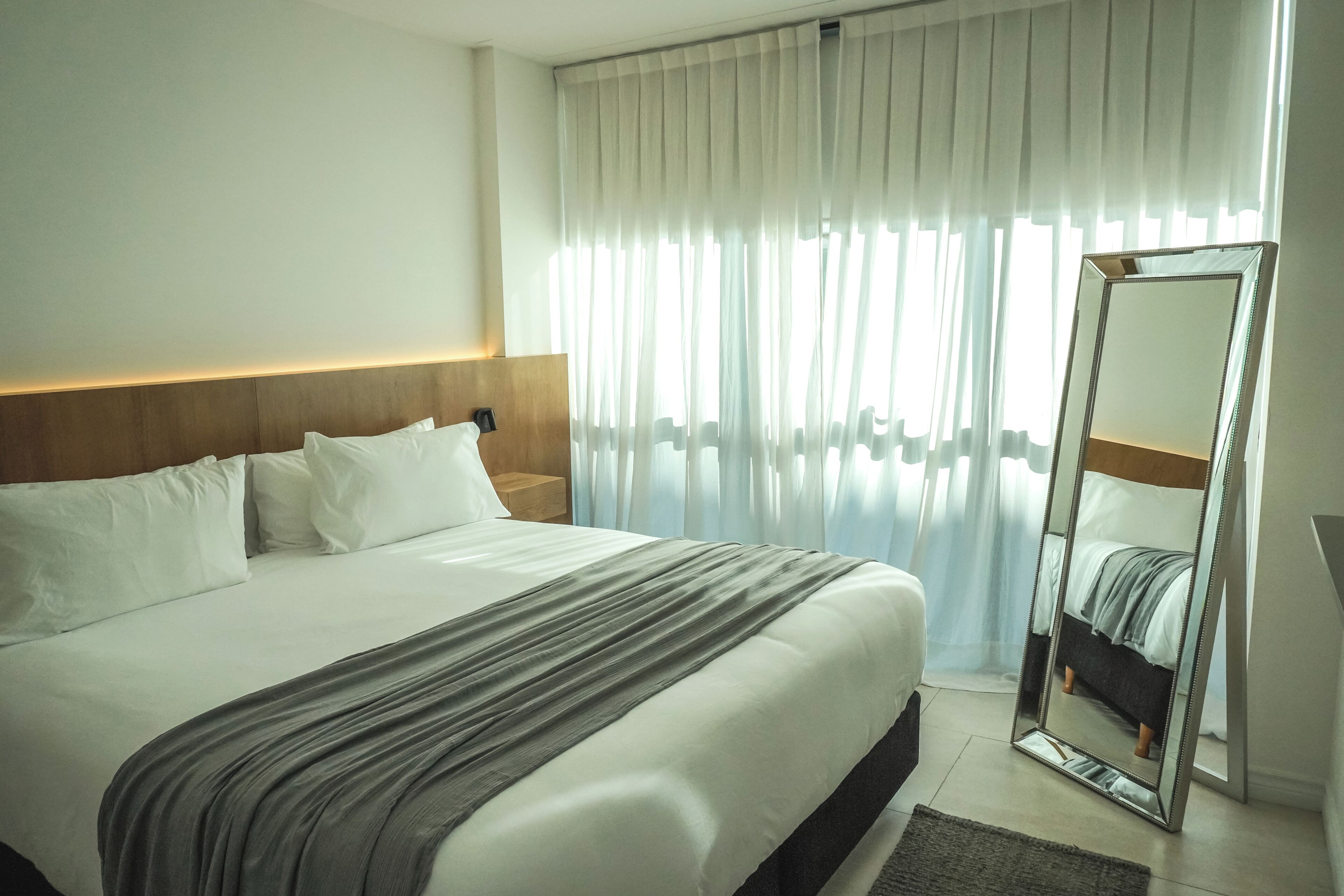 Quarto de hotel com uma cama grande, mesa de cabeceira, espelho e cortinas translúcidas.