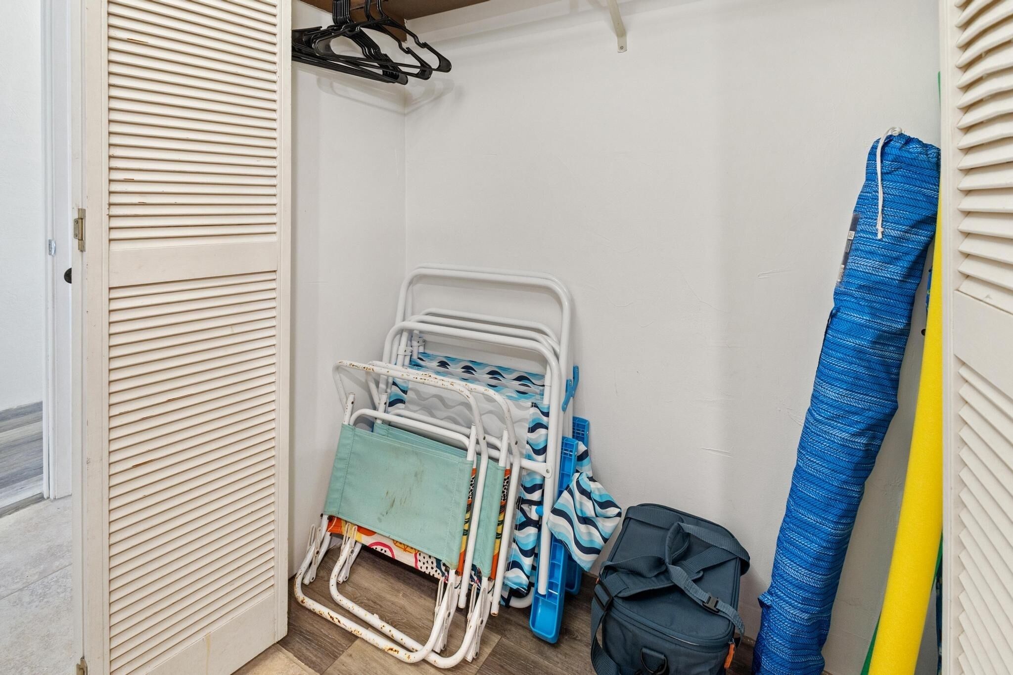Um quarto pequeno com porta fechada, uma cama com cabeceira branca, colchão azul, uma cadeira de metal branca, tapete azul e uma mala preta.