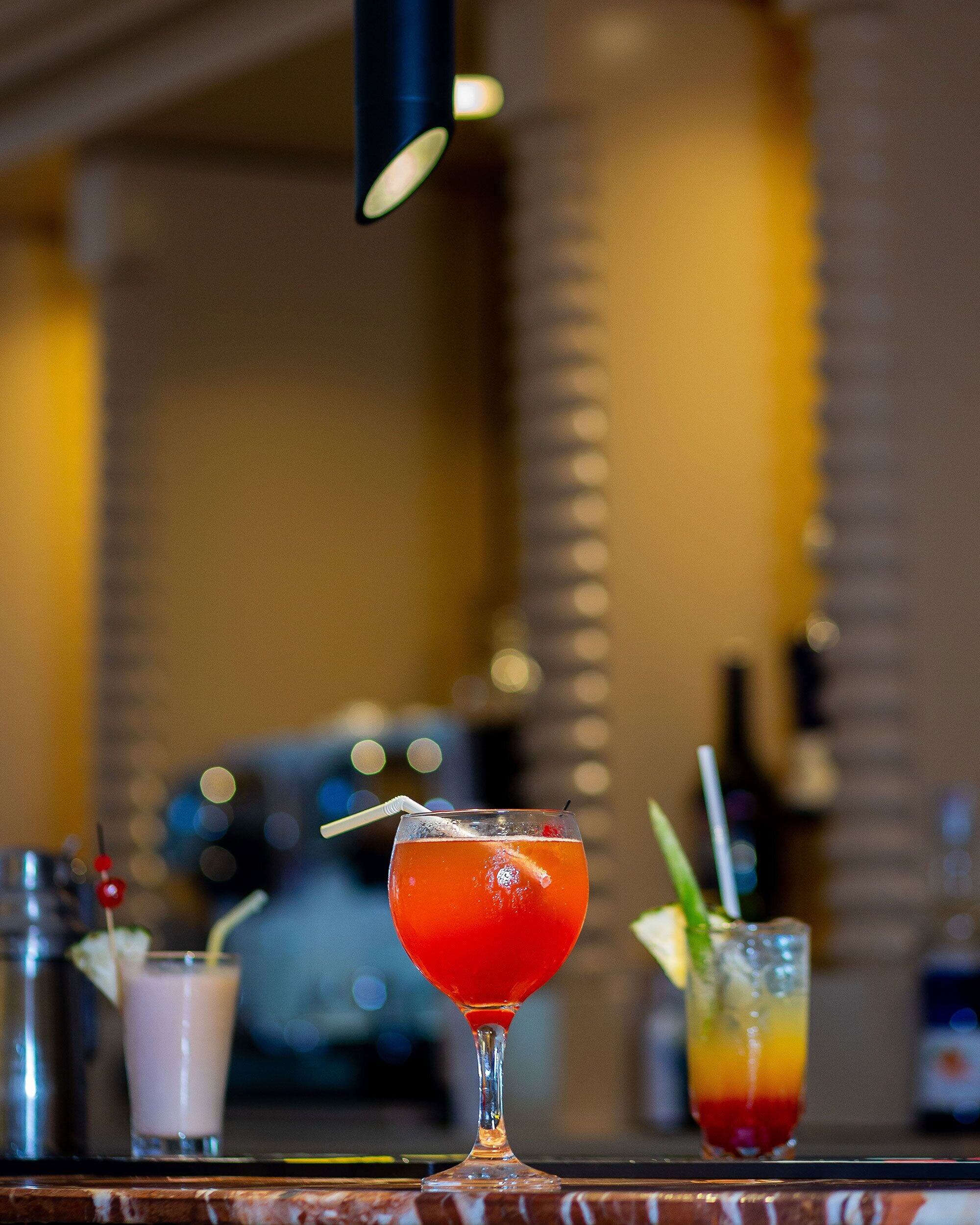 Ambiente de bar com um coquetel vermelho num copo de martini, um copo com canudo e uma fatia de limão, e uma garrafa ao fundo.