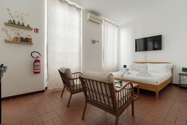 Quarto de hotel com cama, duas cadeiras de vime, uma mesa pequena, televisão e extintor de incêndio na parede.