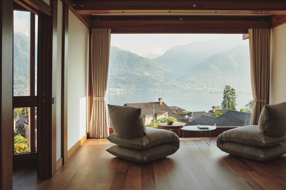 Quarto com uma grande janela, piso de madeira, dois poltronas, uma mesa pequena e vista para montanhas e um lago.