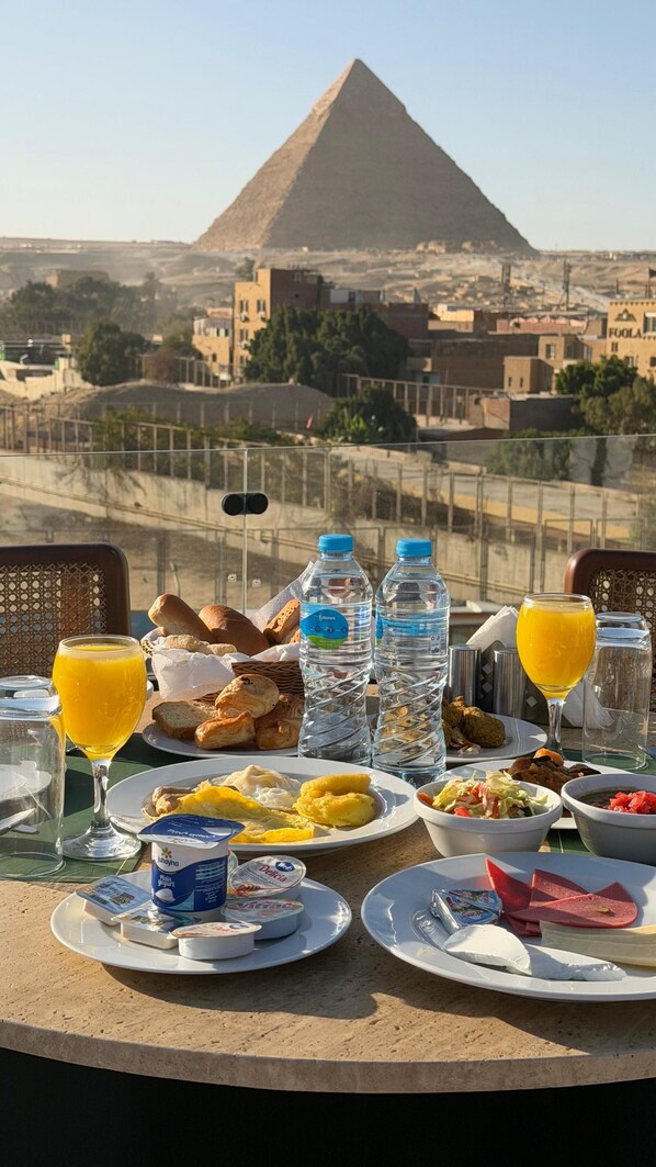 Mesa de café da manhã com diversos alimentos e bebidas, com vista para uma pirâmide e edifícios.