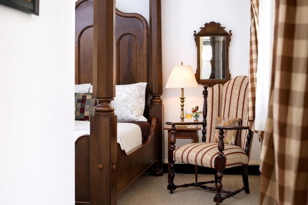 Um quarto com uma cama de dossel de madeira, uma poltrona listrada, uma mesinha de cabeceira com abajur e um espelho na parede.