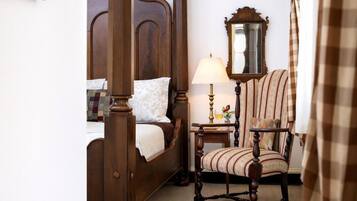 Um quarto com uma cama de dossel de madeira, uma poltrona listrada, uma mesinha de cabeceira com abajur e um espelho na parede.